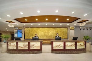 Área de recepção de hotel com um balcão grande, dois recepcionistas e uma parede decorativa.