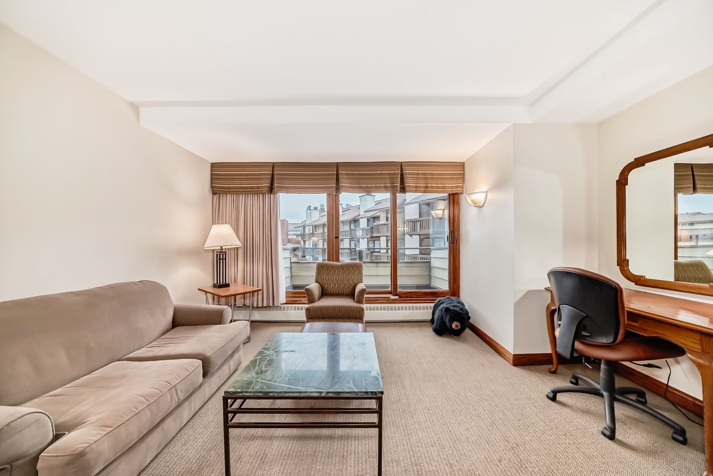 Quarto de hotel com sofá, mesa pequena, cadeira, escrivaninha, luminária, janela grande com cortinas e vista para prédios.
