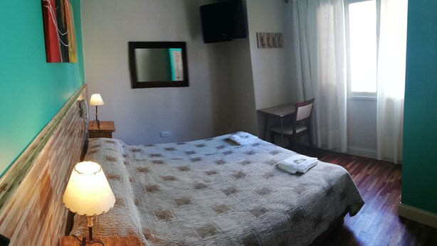 Quarto de hotel com cama, escrivaninha, cadeira, televisão e janela com cortinas.
