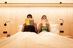 Duas pessoas lendo livros num quarto com paredes de madeira e uma cama com roupa de cama branca.