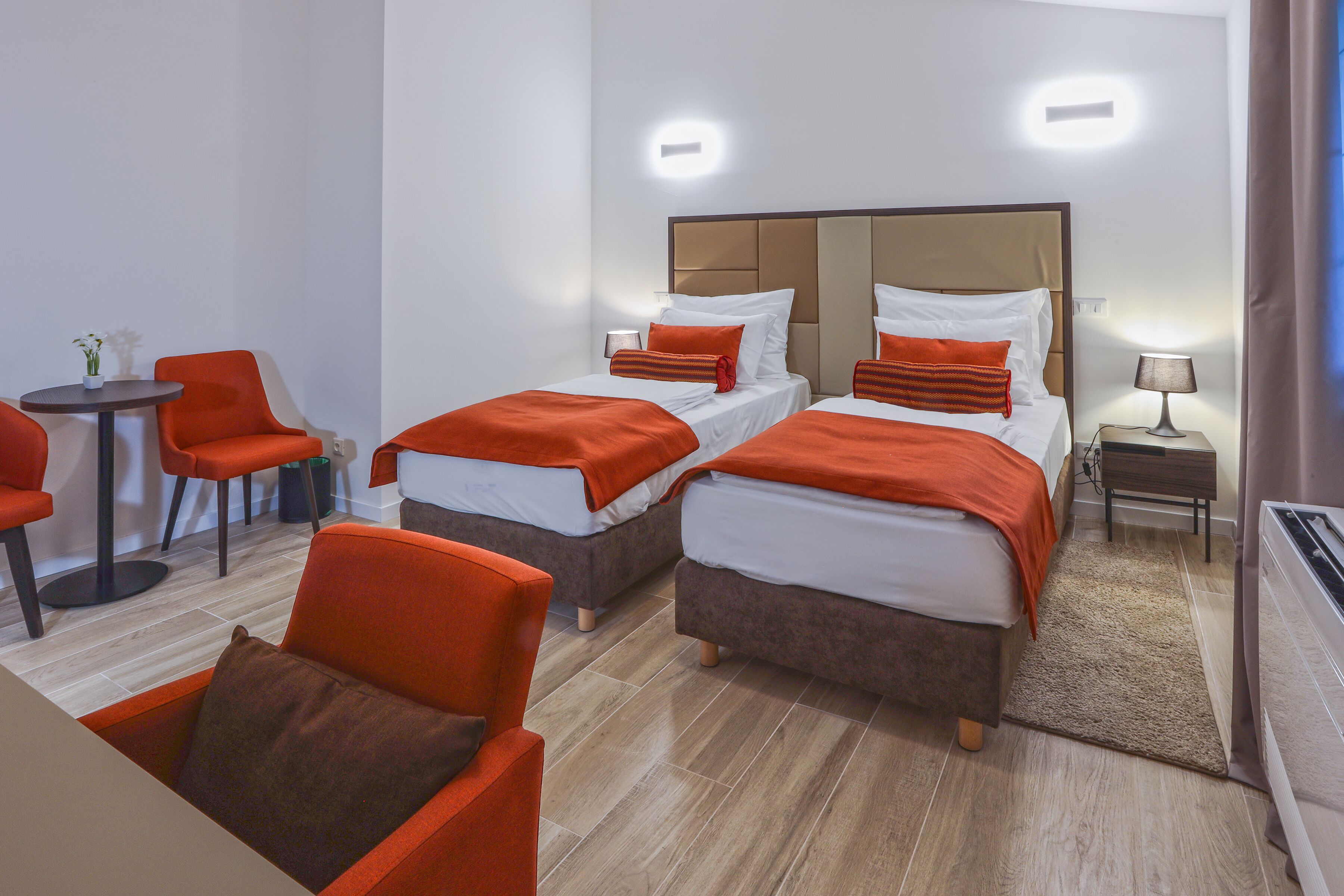 Quarto de hotel com duas camas, cadeiras laranja e marrons, uma pequena mesa e um abajur.