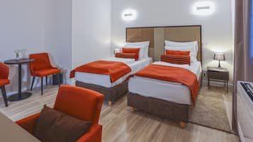 Quarto de hotel com duas camas, cadeiras laranja e marrons, uma pequena mesa e um abajur.