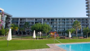 Área de piscina com deck ladrilhado, guarda-sóis e um parquinho ao fundo.