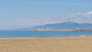 Uma praia arenosa com um molhe rochoso que se estende até o mar, com vários barcos e um farol.