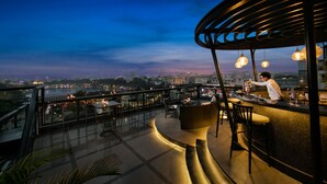 Um bar no terraço com vista panorâmica da cidade, mesas e cadeiras, e um bartender.