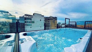 Piscina no terraço com várias banheiras de hidromassagem, cercada por edifícios da cidade e um céu claro.