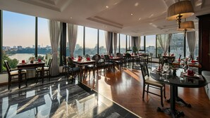 Uma área de jantar com mesas preparadas para uma refeição, grandes janelas com vista para a cidade e um lustre.