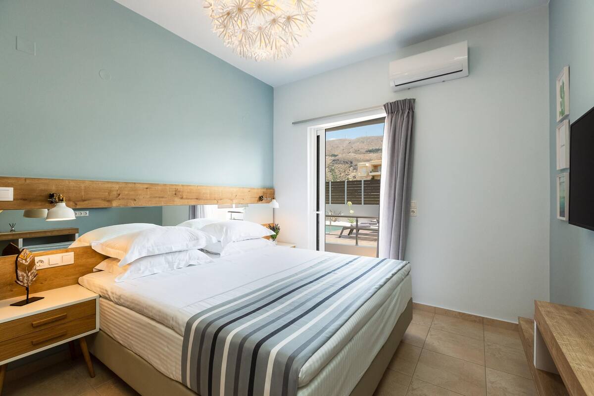 Quarto de hotel com cama, mesa de cabeceira, abajur e vista para o exterior pela janela.