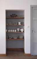 Uma cozinha com prateleiras de madeira que exibem diversos utensílios, uma porta à direita e um interruptor de luz na parede.