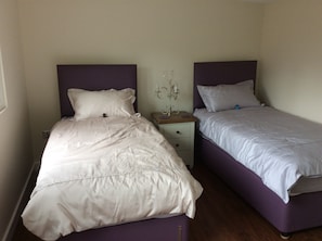 Quarto com duas camas, cada uma com cabeceira, mesa de cabeceira e luminária, além de uma pequena gaveta.
