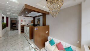 Uma área de recepção de hotel com um balcão de madeira, um candelabro, um relógio e a placa da recepção.