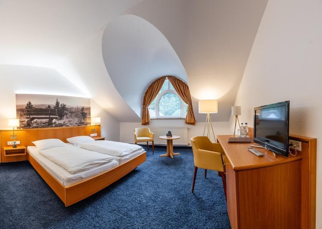Quarto de hotel com cama, escrivaninha, televisão, cadeira, abajur e janela com cortinas.