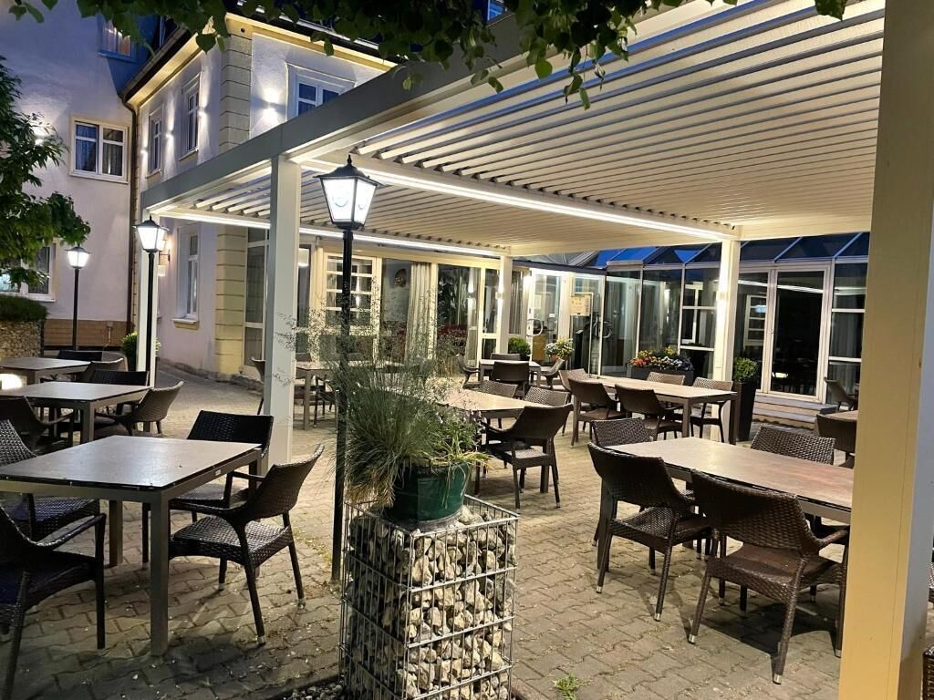 Área de pátio externa com mesas e cadeiras, estrutura de pérgola e um vaso de pedra.