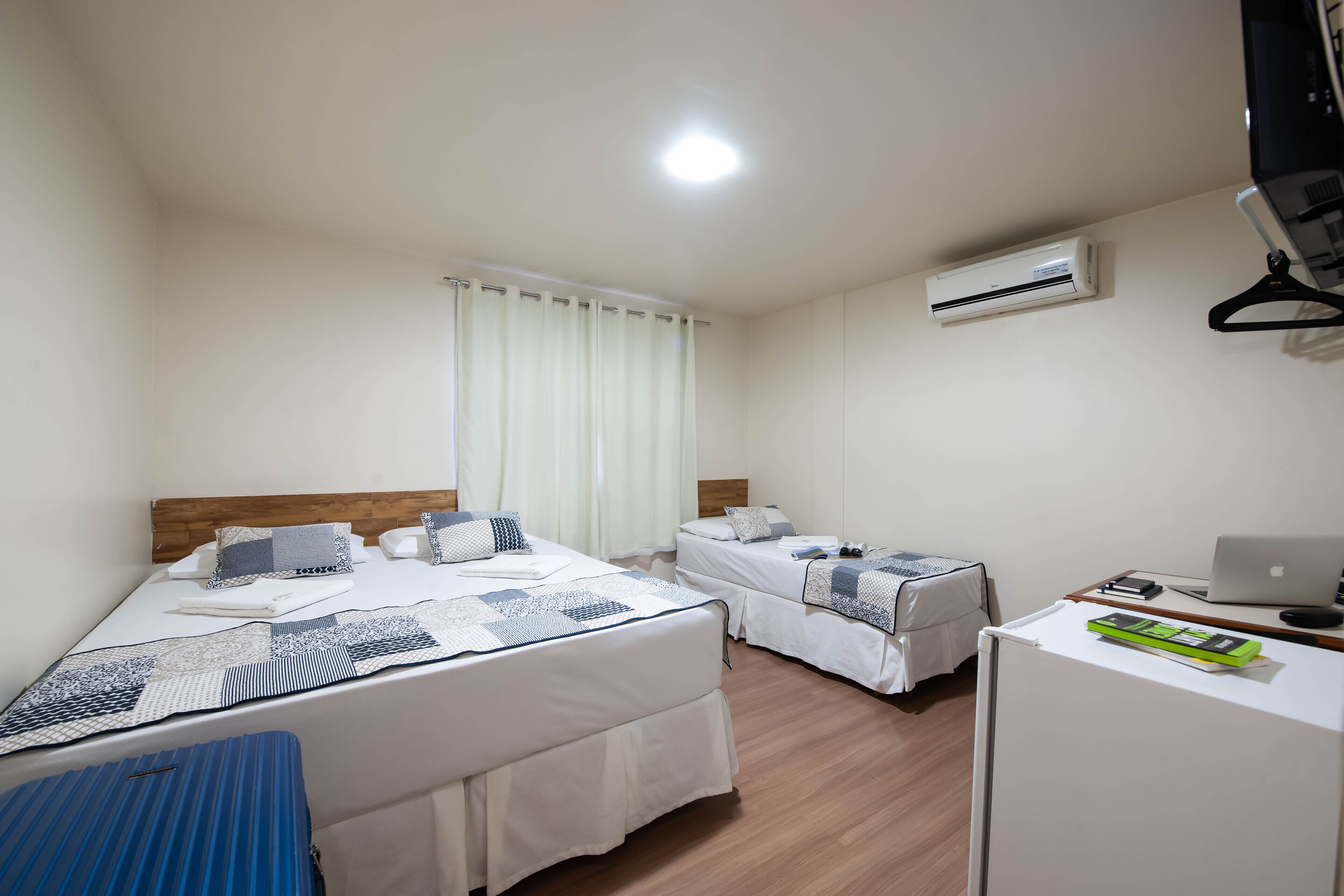 Quarto de hotel com duas camas, uma escrivaninha com um laptop, ar condicionado e um frigorífico.
