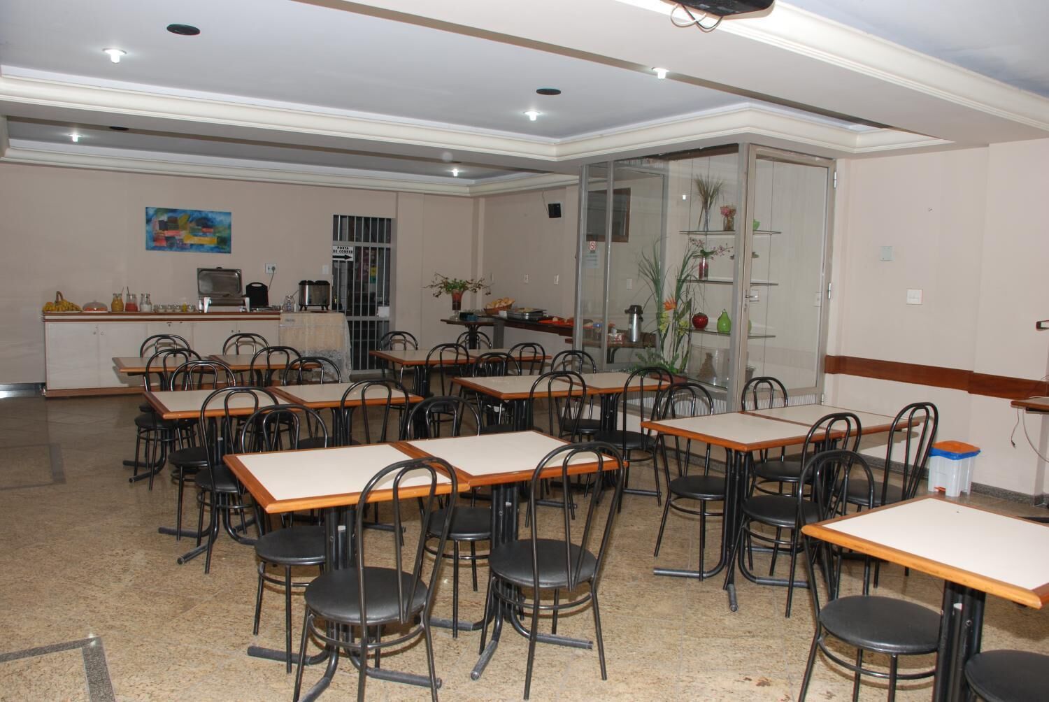Uma área de refeições com várias mesas e cadeiras, uma cozinha com eletrodomésticos e uma planta decorativa.