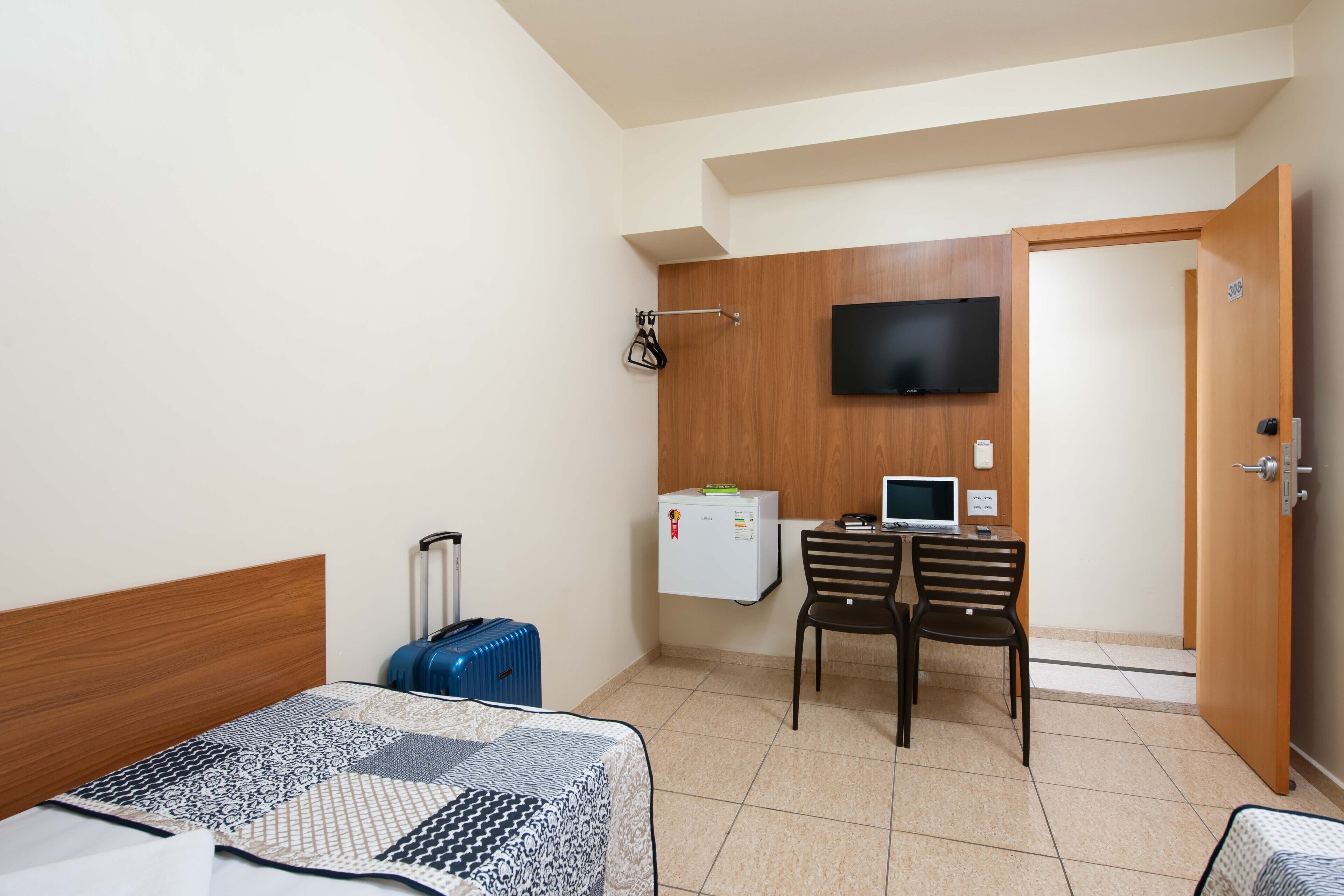 Quarto de hotel com cama, escrivaninha com computador, televisão, mini-geladeira e mala.