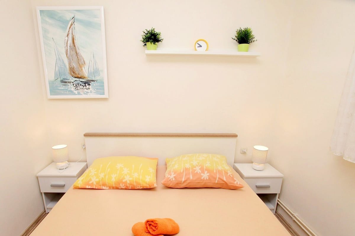 Quarto com cama, duas mesas de cabeceira, prateleira na parede com plantas, relógio e pintura de um veleiro.