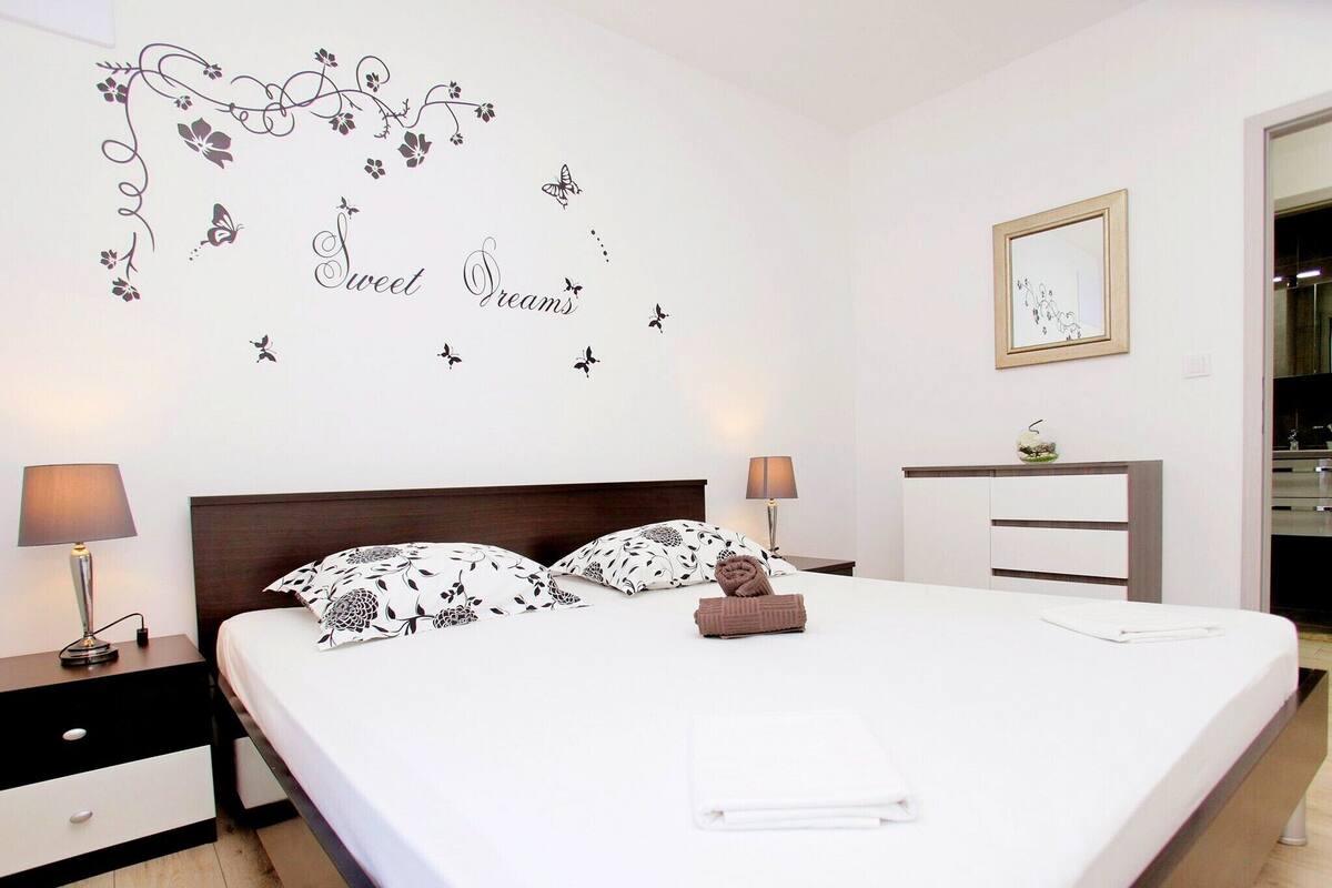Uma cama bem arrumada com almofadas estampadas com flores, uma toalha dobrada e um adesivo de parede com borboletas e o texto “Sonhos Doces”.