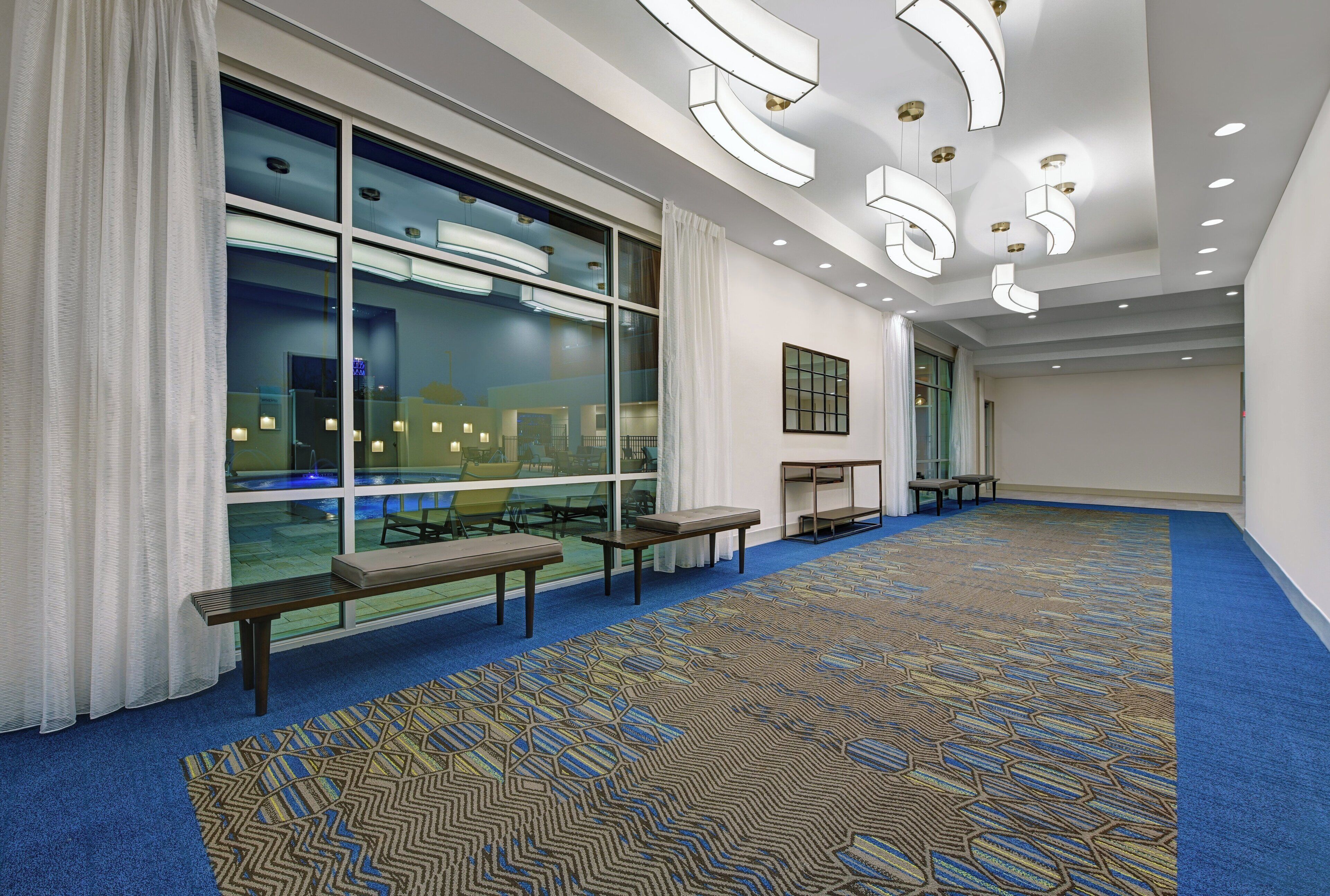 Quarto espaçoso com ampla janela com vista para a área da piscina, carpete estampado e luminária moderna no teto.