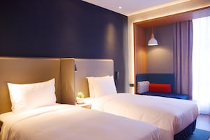Quarto de hotel com duas camas, uma mesa de cabeceira com abajur, uma pequena planta e uma janela com cortinas.