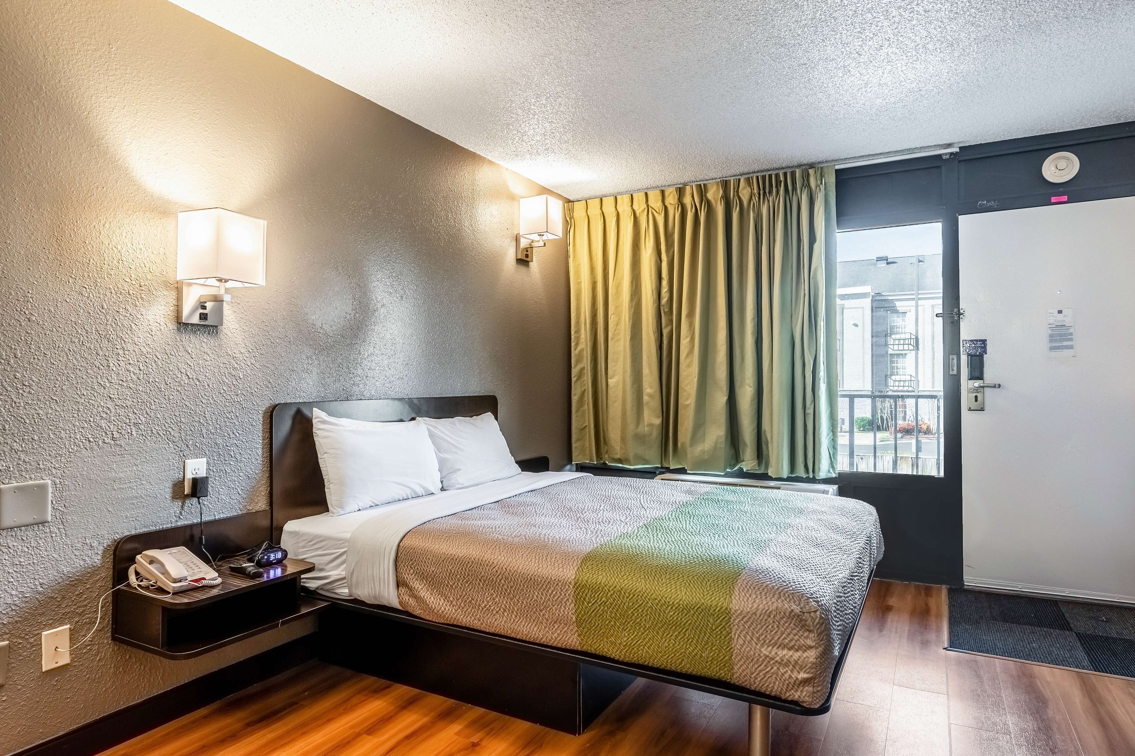 Quarto de hotel com cama, mesa de cabeceira com telefone e carregador, luminária na parede e janela com cortinas.