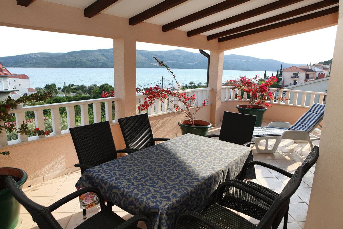 Uma varanda com mesa e cadeiras, plantas em vasos e vista para o mar e montanhas.