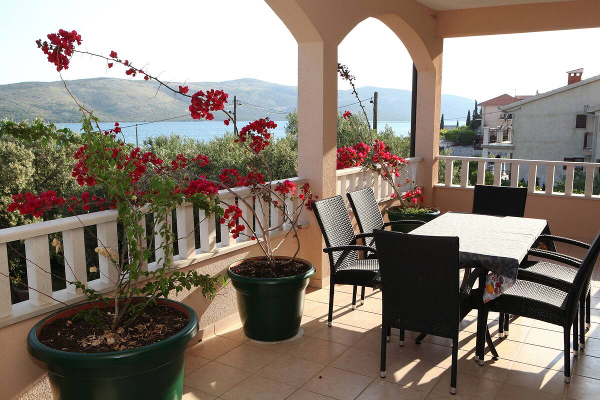 Varanda com mesa e cadeiras, plantas em vasos e vista para o mar e montanhas.