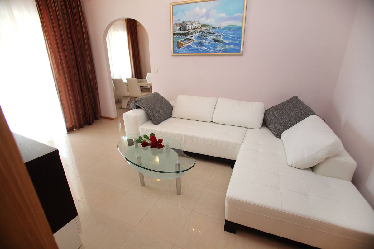 Uma sala de estar com um sofá branco, uma mesa de centro de vidro com flores e um quadro de barcos na parede.