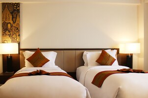 Quarto de hotel com duas camas, cada uma com roupa de cama branca e almofadas decorativas castanhas. Há duas luminárias de cabeceira e uma tapeçaria grande na parede.