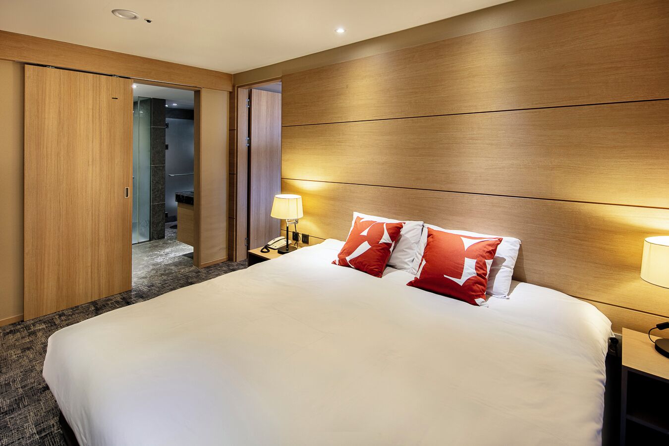 Quarto de hotel com uma cama grande, cabeceira de madeira, uma mesa de cabeceira com abajur e um telefone.