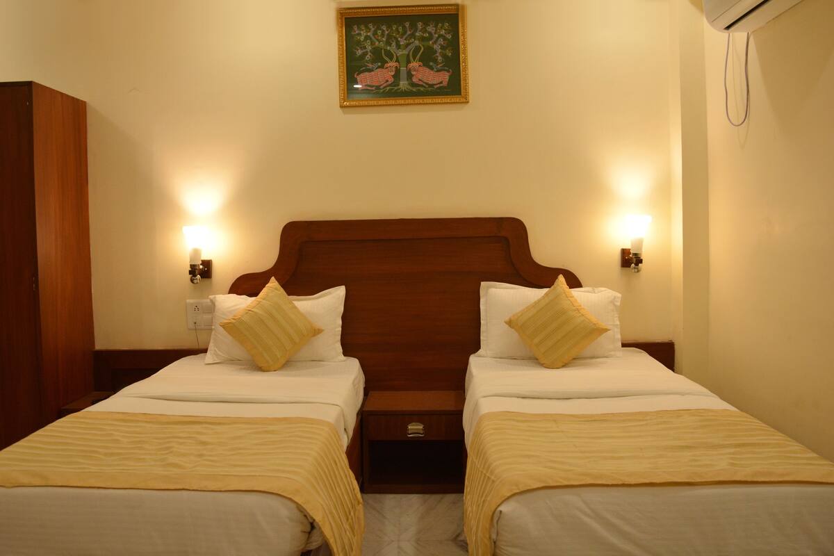 Quarto duplo com duas camas, cada uma com cabeceira de madeira, roupa de cama branca e duas almofadas. Existem luminárias fixadas na parede, de cada lado da cama.