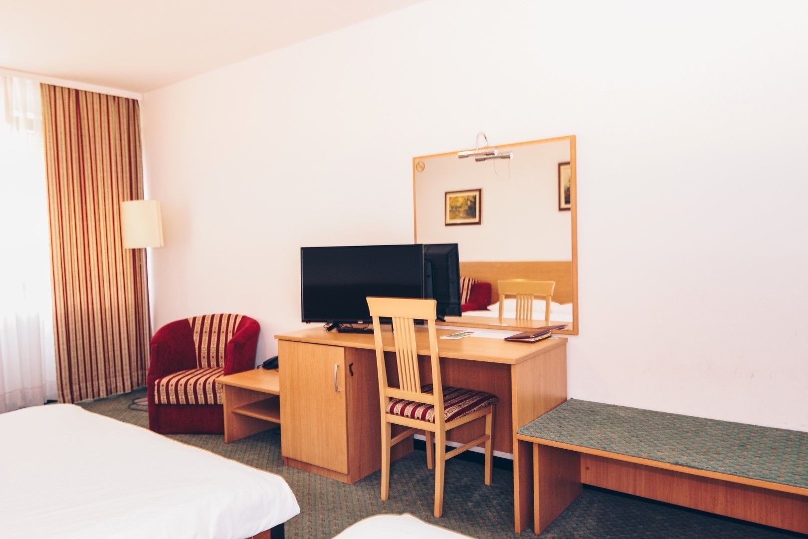 Quarto de hotel com cama, escrivaninha com computador, cadeira, televisão e espelho.