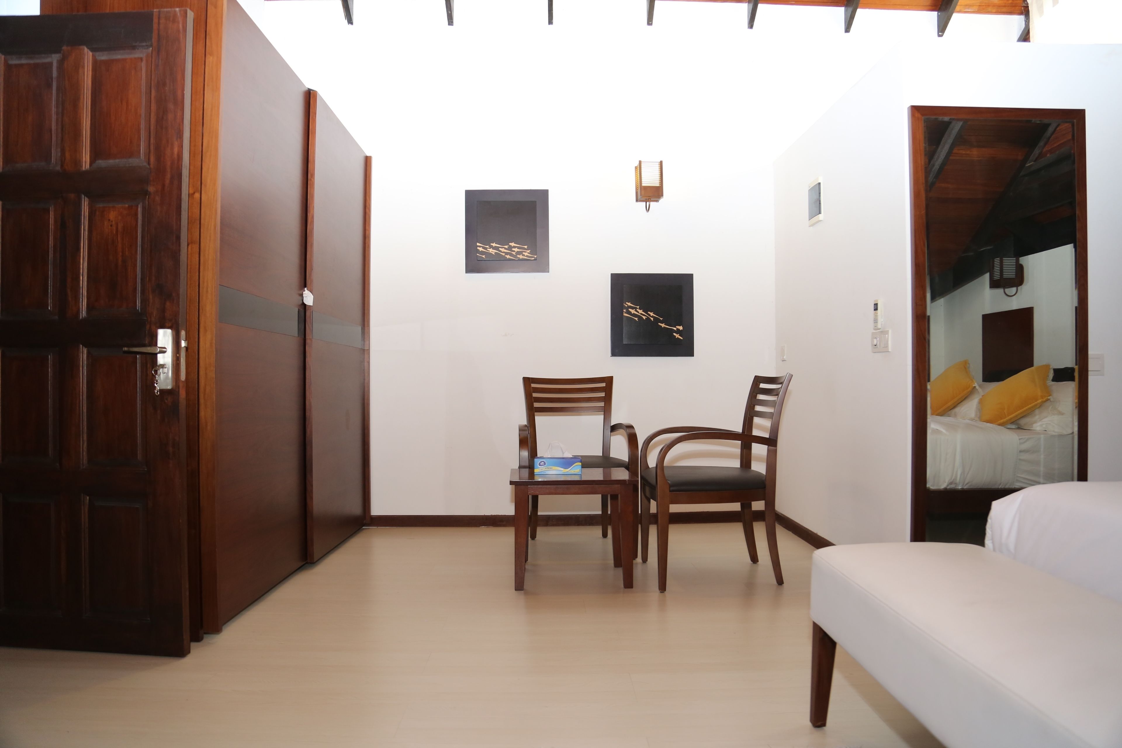 Um quarto com porta de madeira, duas cadeiras, uma mesinha, uma cama e duas imagens emolduradas na parede.