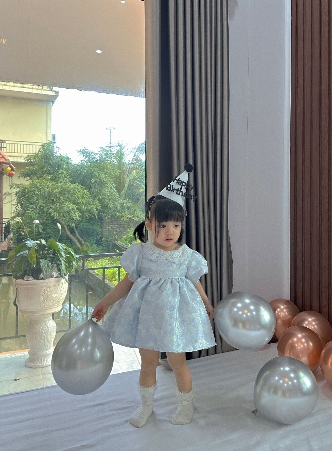 Criança de vestido branco e chapéu de festa segurando um balão, em ambiente interno com vista para uma varanda.