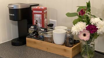 Uma bancada de cozinha com uma cafeteira, um recipiente em formato de cabine telefônica vermelha, uma bandeja com sachês de café, canecas e um vaso com flores.