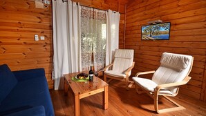 Interior de uma cabana de madeira com um sofá azul, duas cadeiras brancas estofadas, uma mesa de madeira com uma garrafa de vinho e copos, e um quadro na parede.