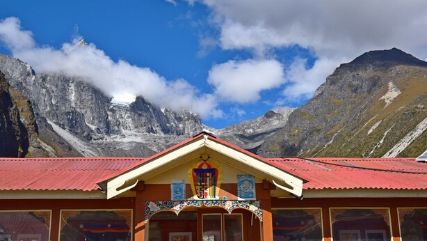 Um chalé de montanha com telhado vermelho e arquitetura tradicional, com as montanhas rochosas e o céu azul claro ao fundo.