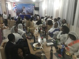 Um salão com mesas redondas, algumas com comida e bebidas, e um palco com um cenário ao fundo.