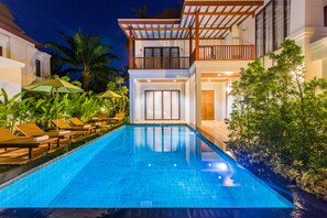 Uma piscina com espreguiçadeiras e guarda-sóis, rodeada por plantas tropicais e um edifício de dois andares com varandas.