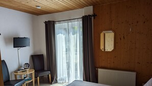 Quarto de hotel com uma cama, uma cadeira, uma pequena mesa com uma chaleira, uma televisão e uma janela com cortinas.