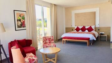 Quarto de hotel com cama, sofá, poltrona, mesinha e um quadro na parede.