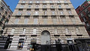 Um edifício de vários andares com arquitetura clássica, destacando uma entrada central com arco decorativo, várias varandas e uma loja no térreo com sinalização.