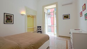 Quarto com cama, guarda-roupa, cadeira e escrivaninha. Há uma janela e uma porta que dá para uma varanda.