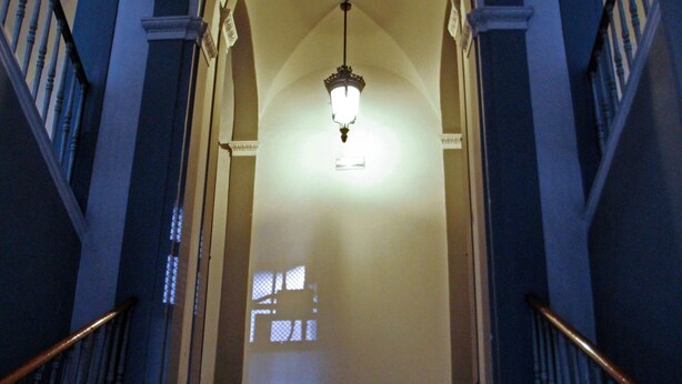 Uma escadaria imponente com um candelabro, ladeada por colunas, que leva a um andar superior.