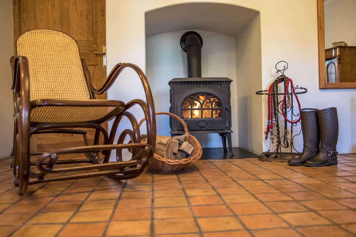 Um quarto com uma cadeira de madeira, uma cadeira de fibra, uma lareira com lenha e um porta-roupas com botas.