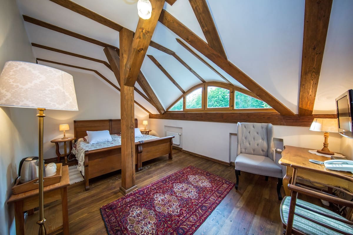 Quarto com teto de madeira, uma janela grande, cama, cadeira, escrivaninha e abajur.
