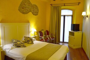 Quarto de hotel com uma cama grande, duas cadeiras, uma televisão e janela com cortinas.