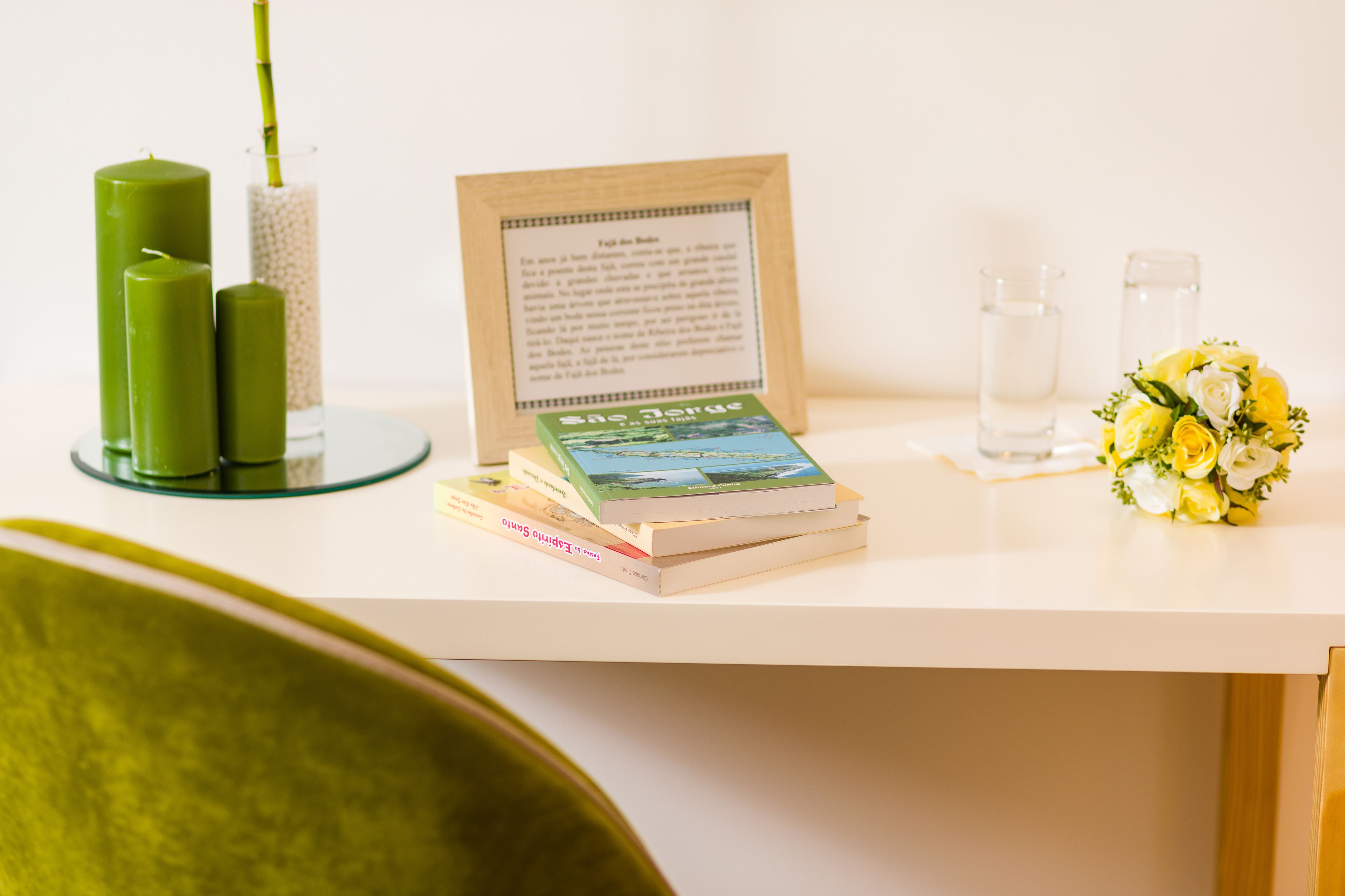 Uma mesa branca com uma pilha de livros, um texto emoldurado, um copo de água e um buquê de flores amarelas.