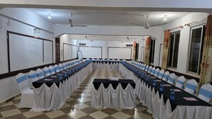 Uma sala de conferências com uma mesa comprida, cadeiras com fitas azuis, um projetor e ventiladores de teto.