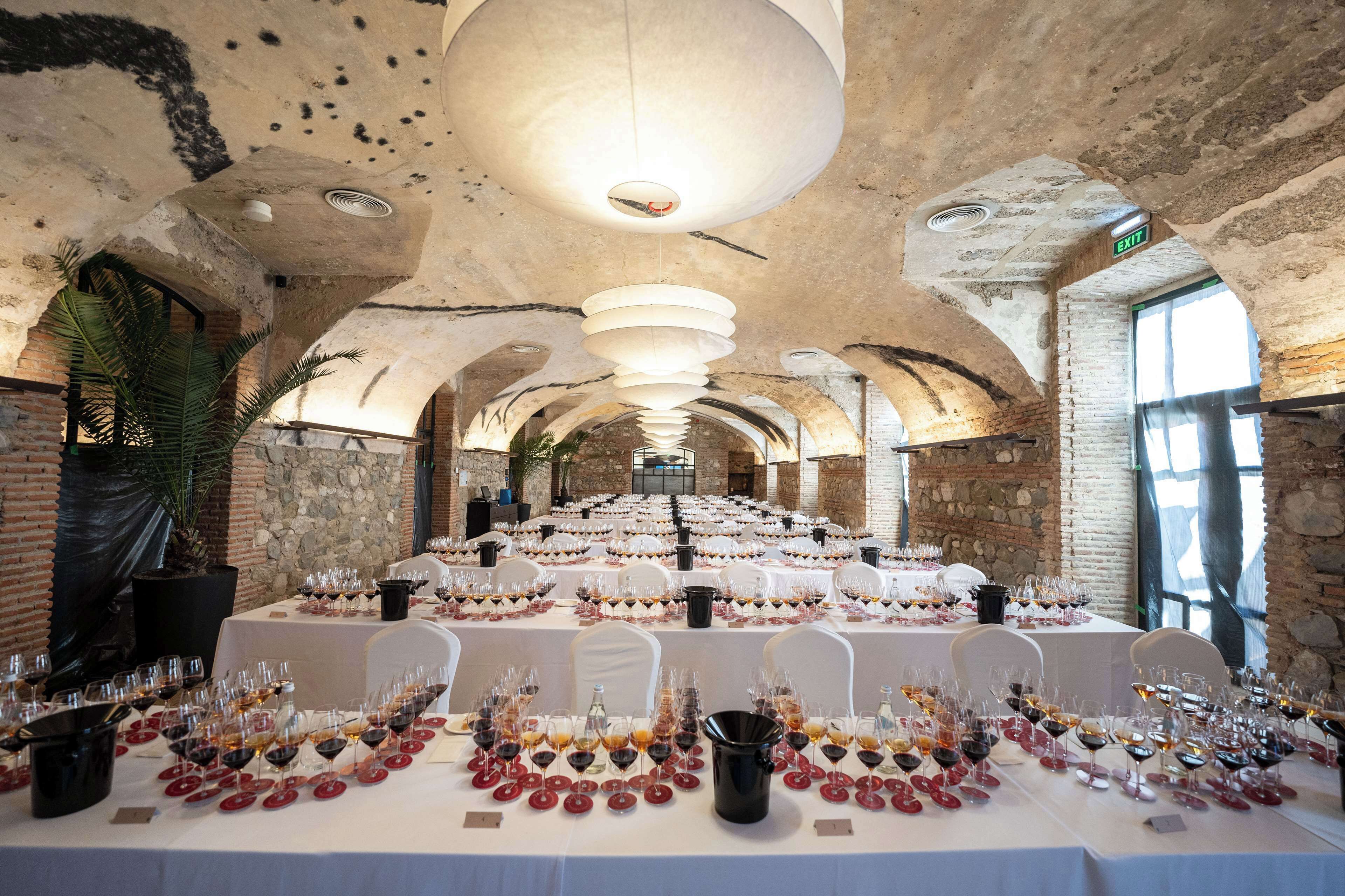 Uma sala de banquetes com várias mesas dispostas para um evento formal, com toalhas brancas, copos de vinho e cadeiras.